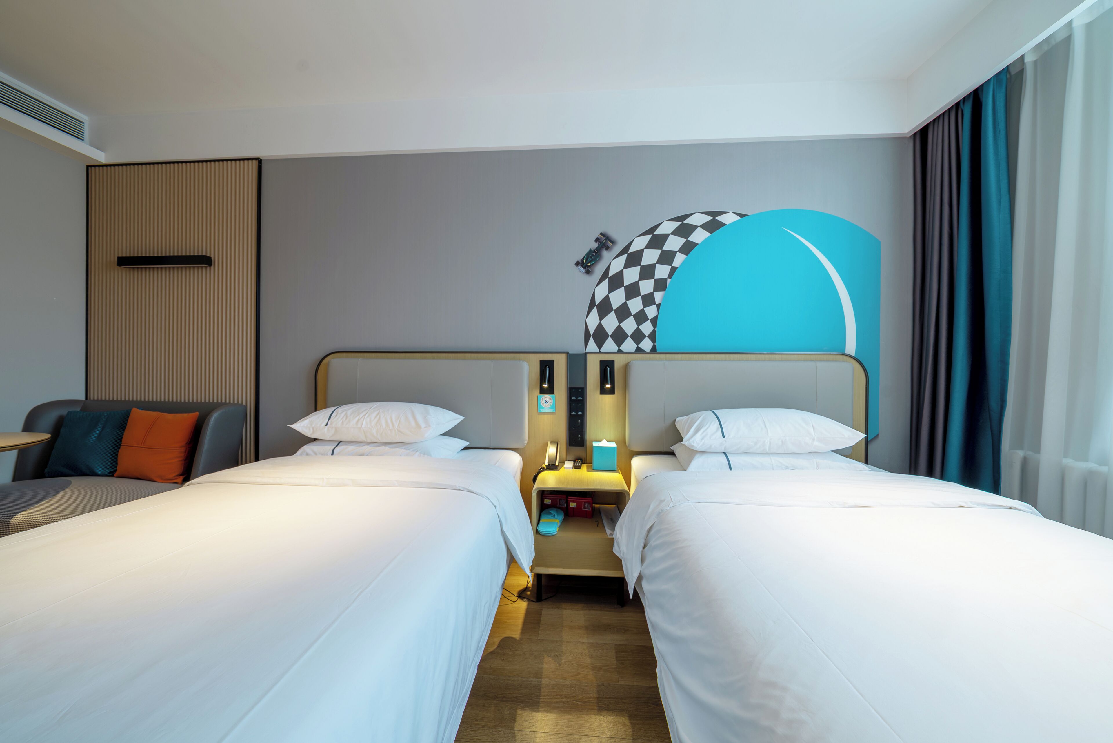 Quarto de hotel com duas camas, uma escrivaninha, uma cadeira, uma mesinha, um espelho e uma janela com cortinas.