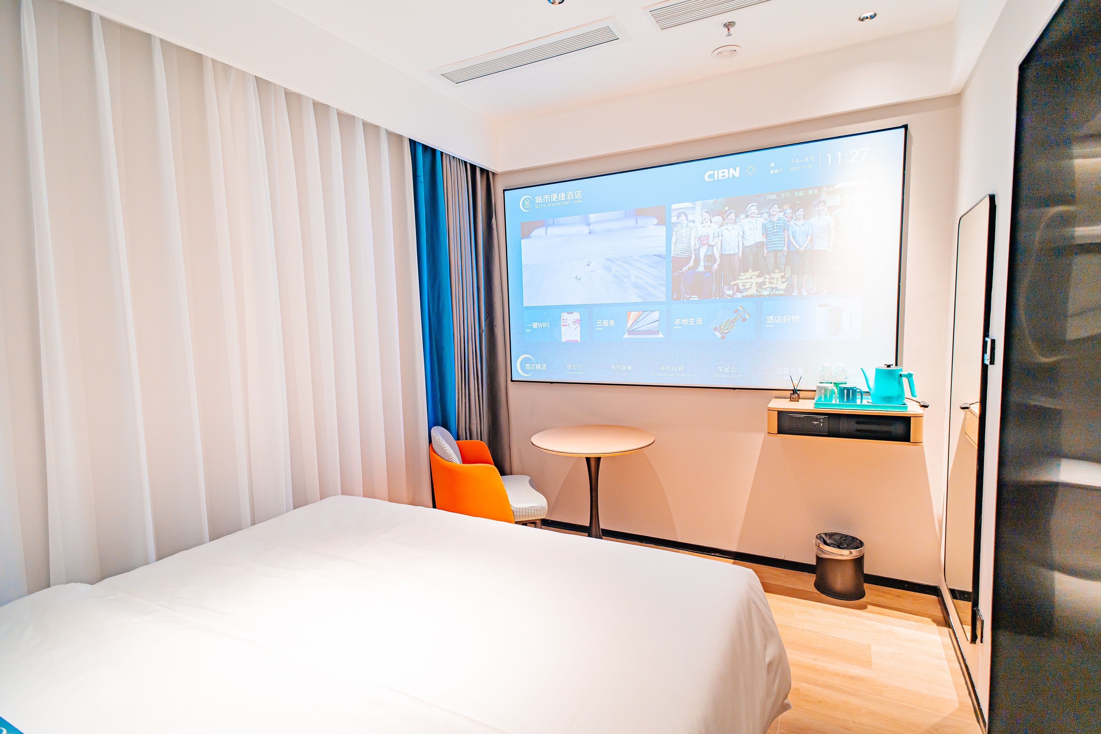 Quarto de hotel com cama, uma pequena mesa, tela de projetor e televisão.