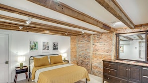 Quarto com cama, mesas de cabeceira, cômoda, parede de tijolos e teto com vigas aparentes.