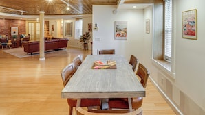 Uma sala de jantar ampla, com mesa de madeira e cadeiras de couro, rodeada por uma parede de tijolos e uma grande janela.