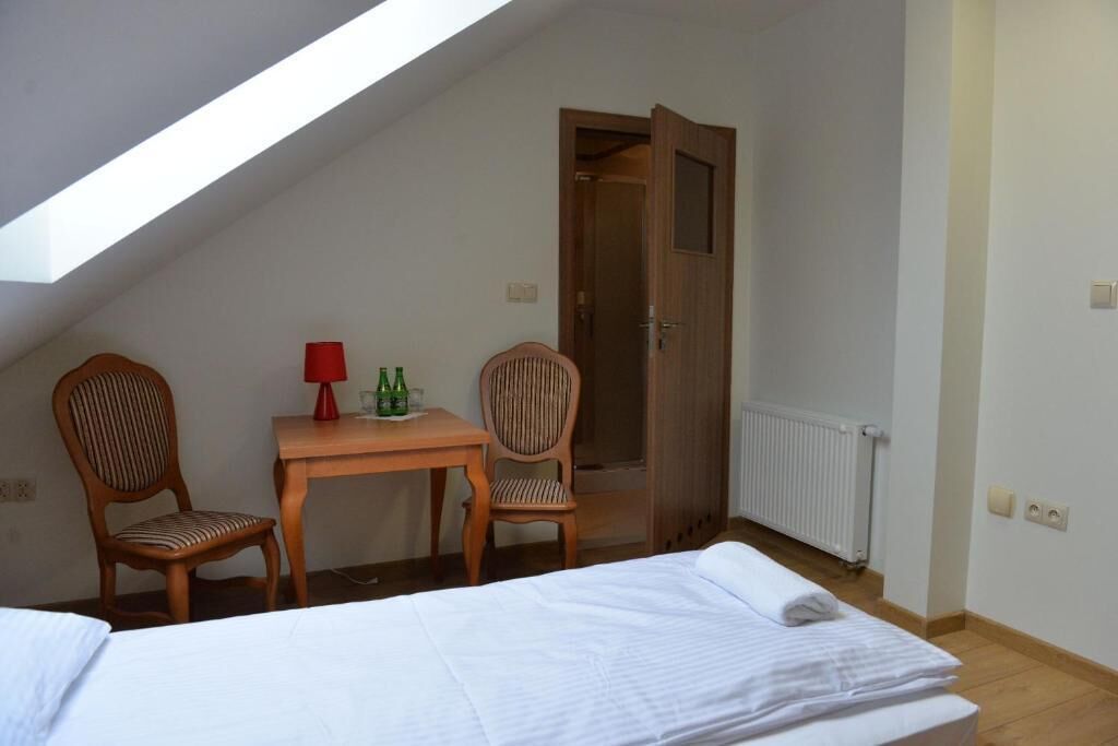 Um quarto pequeno e arrumado com teto inclinado, uma cama, duas cadeiras de madeira, uma mesa pequena com abajur e uma porta.