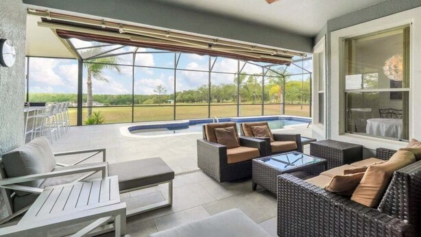Uma área de pátio externa com móveis de vime, ventilador de teto e vista para uma piscina e vegetação.