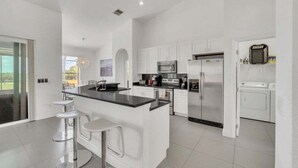 Uma cozinha moderna com armários brancos, uma ilha central com banquetas, eletrodomésticos de aço inoxidável e uma área de lavanderia.