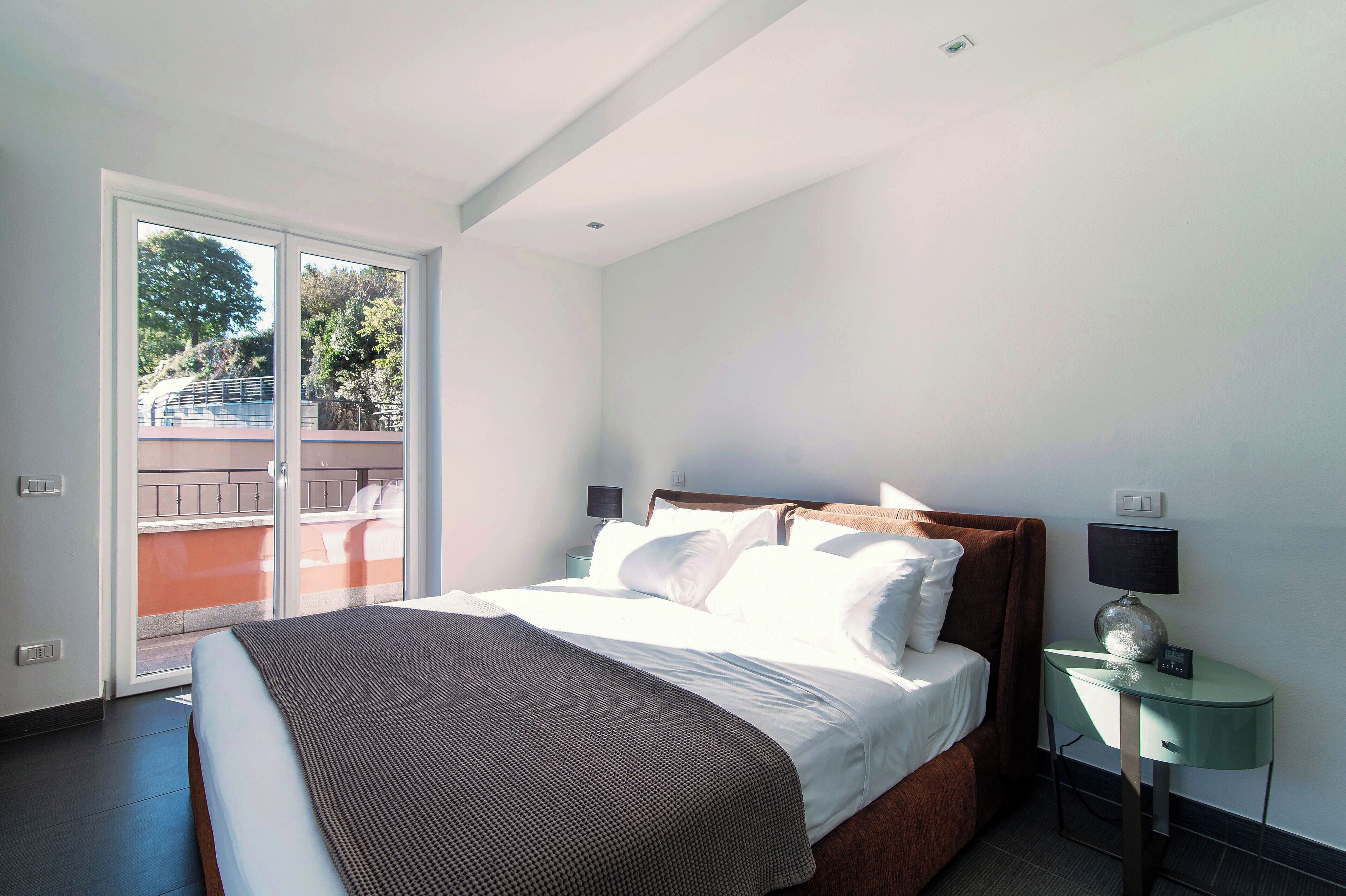 Quarto de hotel com cama, mesa de cabeceira, luminária e porta de vidro deslizante para área externa.