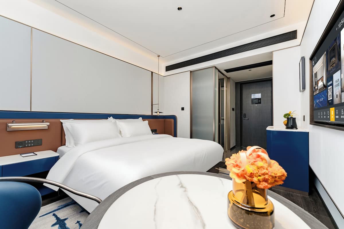 Quarto de hotel com cama, mesa de cabeceira e pequena escrivaninha com um vaso de flores.
