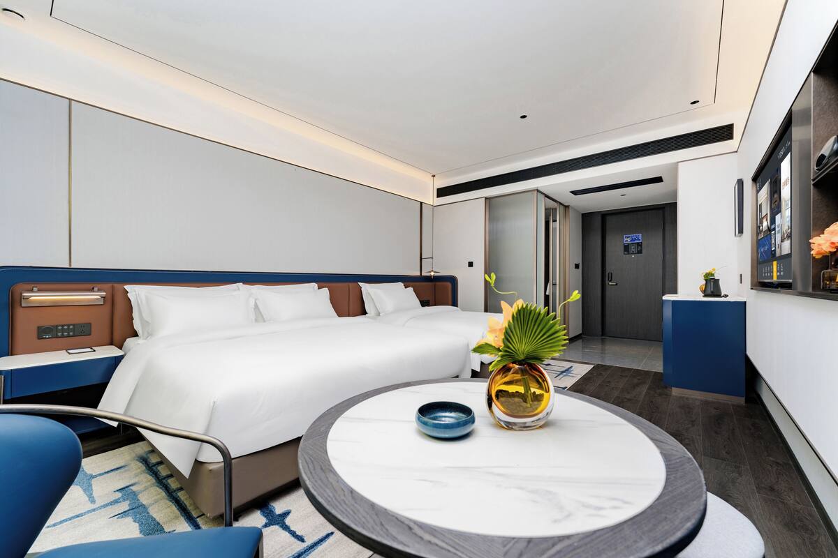 Quarto de hotel moderno com cama, mesa de cabeceira, mesa redonda e um pequeno vaso com flores.