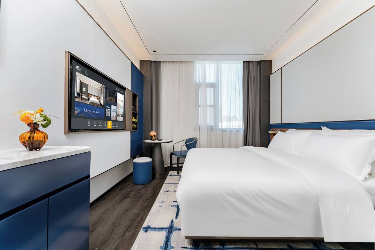 Quarto de hotel com cama, escrivaninha, cadeira, televisão e um vaso com flores.