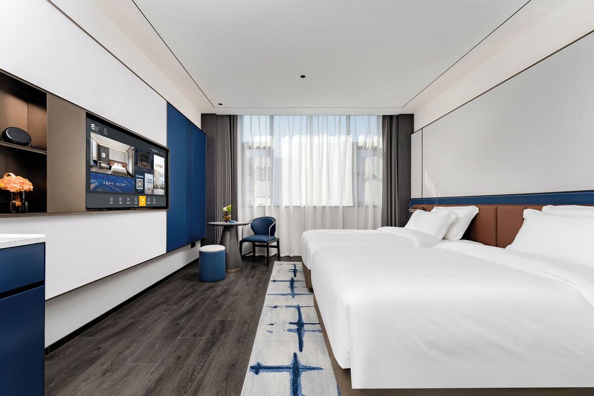 Quarto de hotel moderno com uma cama grande, uma escrivaninha com computador, televisão e vista para a cidade através de grandes janelas.