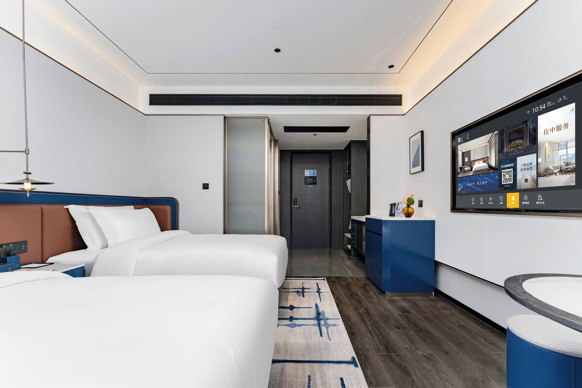 Quarto de hotel moderno com cama, mesa de cabeceira e um painel digital na parede.