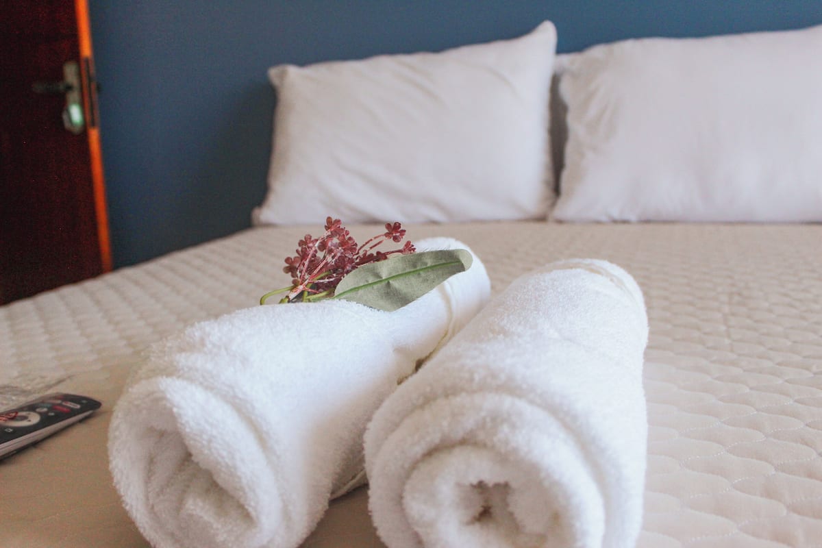 Cama bem arrumada com lençóis brancos, um controle remoto e uma flor decorativa no travesseiro.