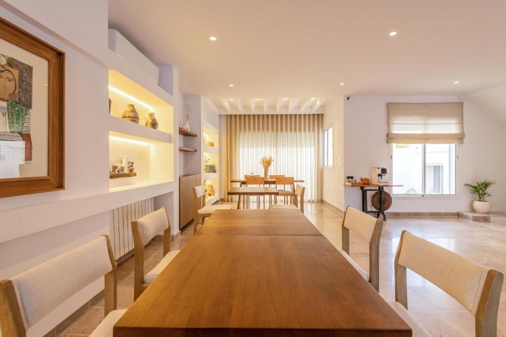 Uma sala de jantar espaçosa com uma longa mesa de madeira e cadeiras, área de cozinha com fogão e armários, e uma grande janela com cortinas.