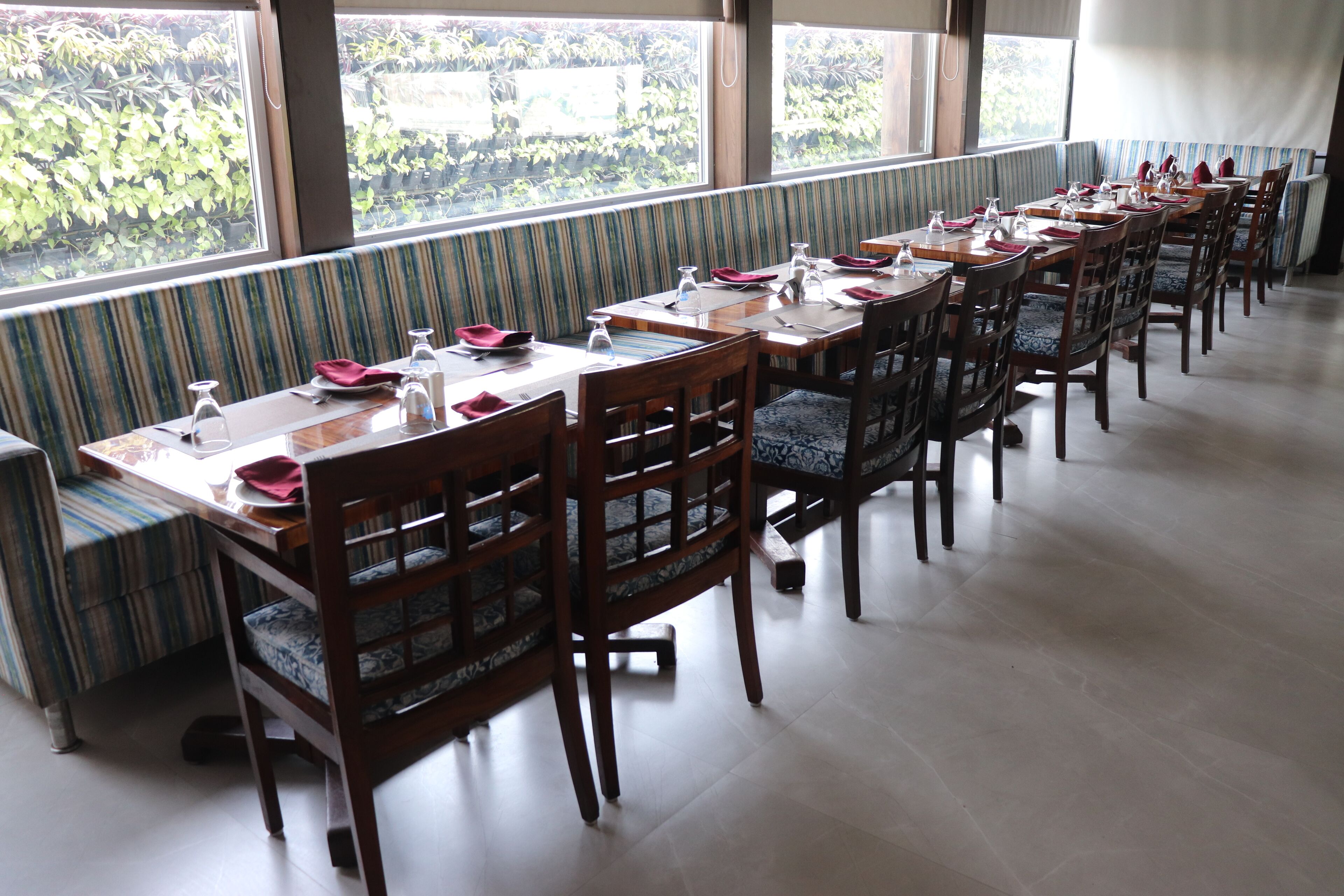 Um restaurante com mesas e cadeiras compridas, grandes janelas com vista para o verde e um banco listrado ao longo da parede.