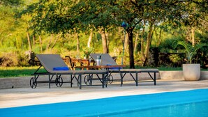 Área à beira da piscina com espreguiçadeiras e uma mesa, cercada por vegetação.