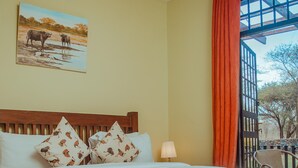 Uma cama com estrutura de madeira, almofada com estampas de animais, abajur e vista para árvores através de uma janela com cortinas laranja.