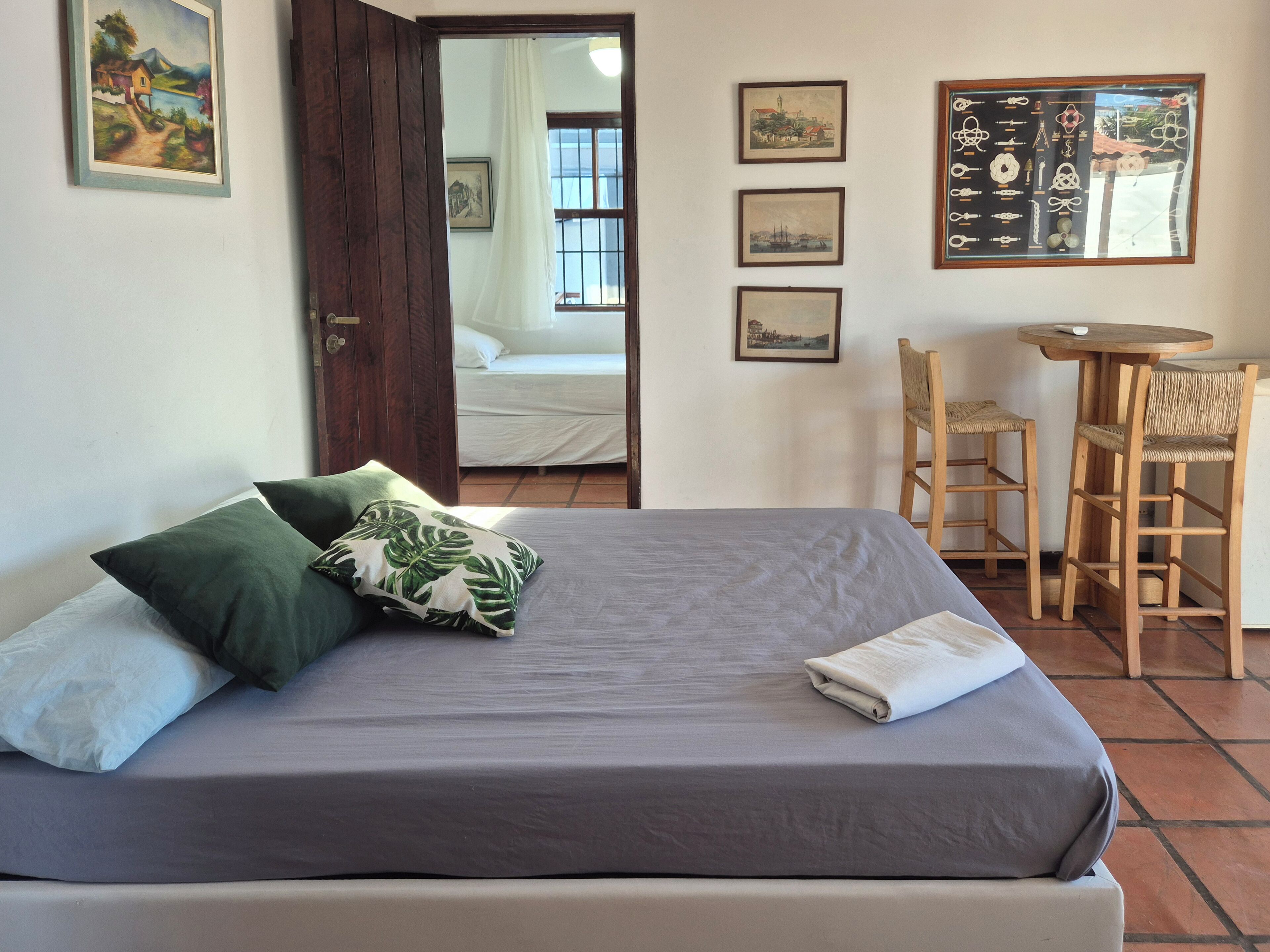 Cama bem arrumada com colcha cinza e duas almofadas, porta de madeira que dá para um quarto com cama, e uma pequena mesa de madeira com duas cadeiras.
