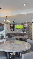 Uma área de bar moderna com uma televisão de tela plana exibindo um evento esportivo, um armário para vinhos e um espaço de estar com mesas e cadeiras redondas.