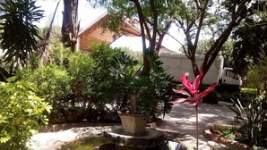 Um jardim com uma fonte, diversas plantas e um veículo branco ao fundo.