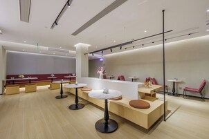 Uma área de convívio moderna e bem iluminada, com diversas opções de assento incluindo mesas e bancos, um balcão de recepção e um vaso decorativo com flores.
