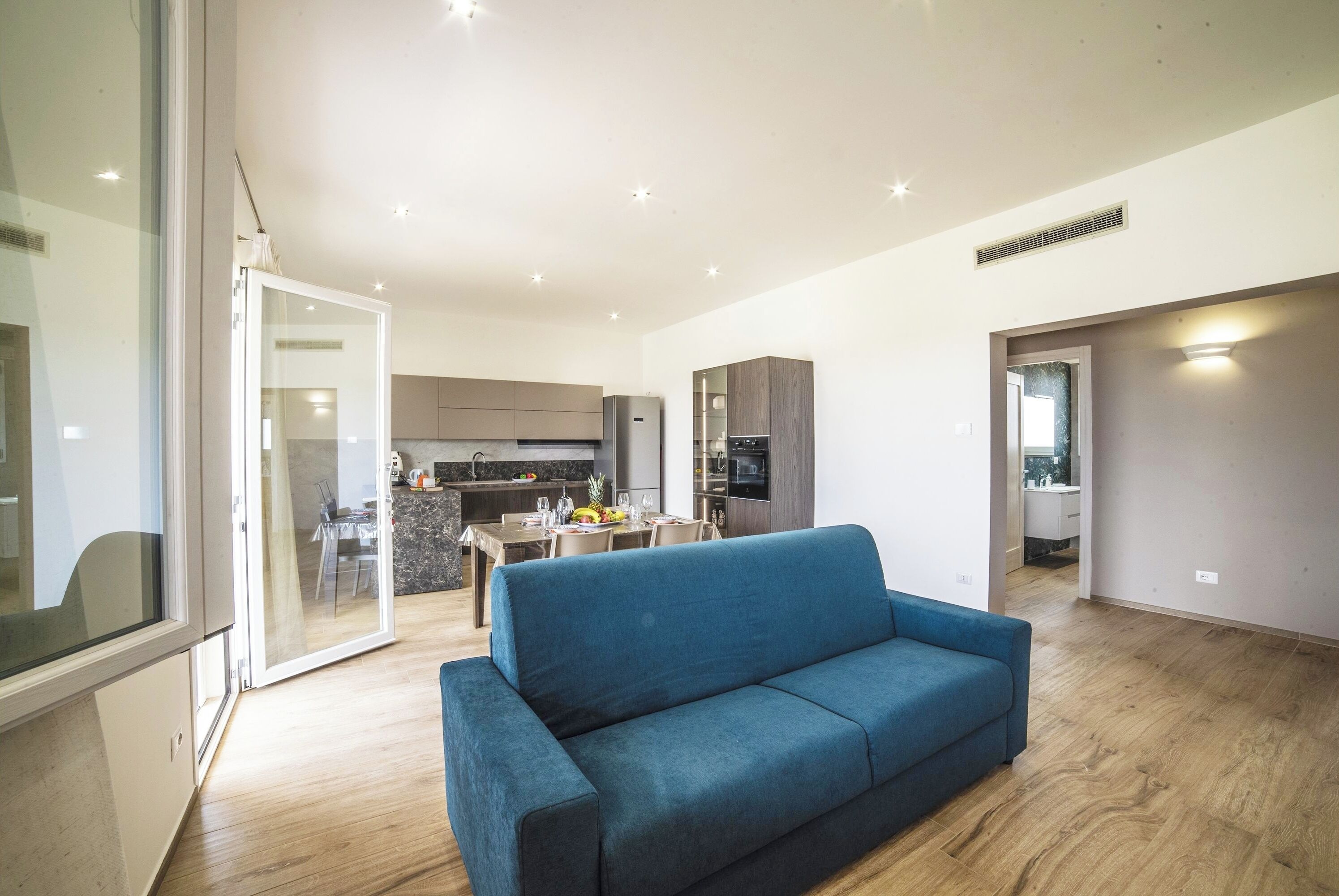 Uma sala de estar moderna com um sofá azul, uma área de jantar com mesa e cadeiras, e uma cozinha ao fundo.