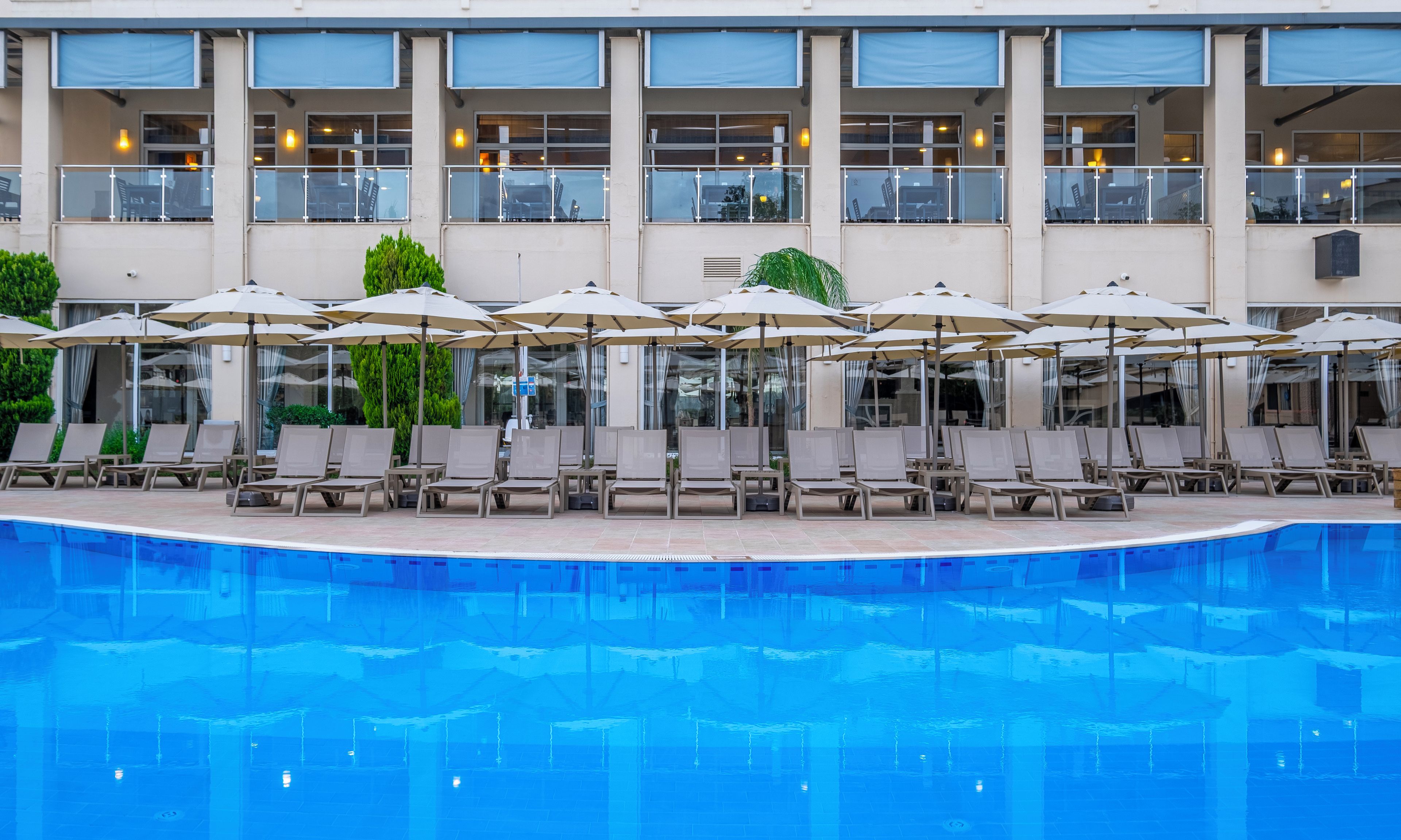 Piscina com espreguiçadeiras e guarda-sóis, um edifício com vários andares e janelas amplas, e um céu claro.