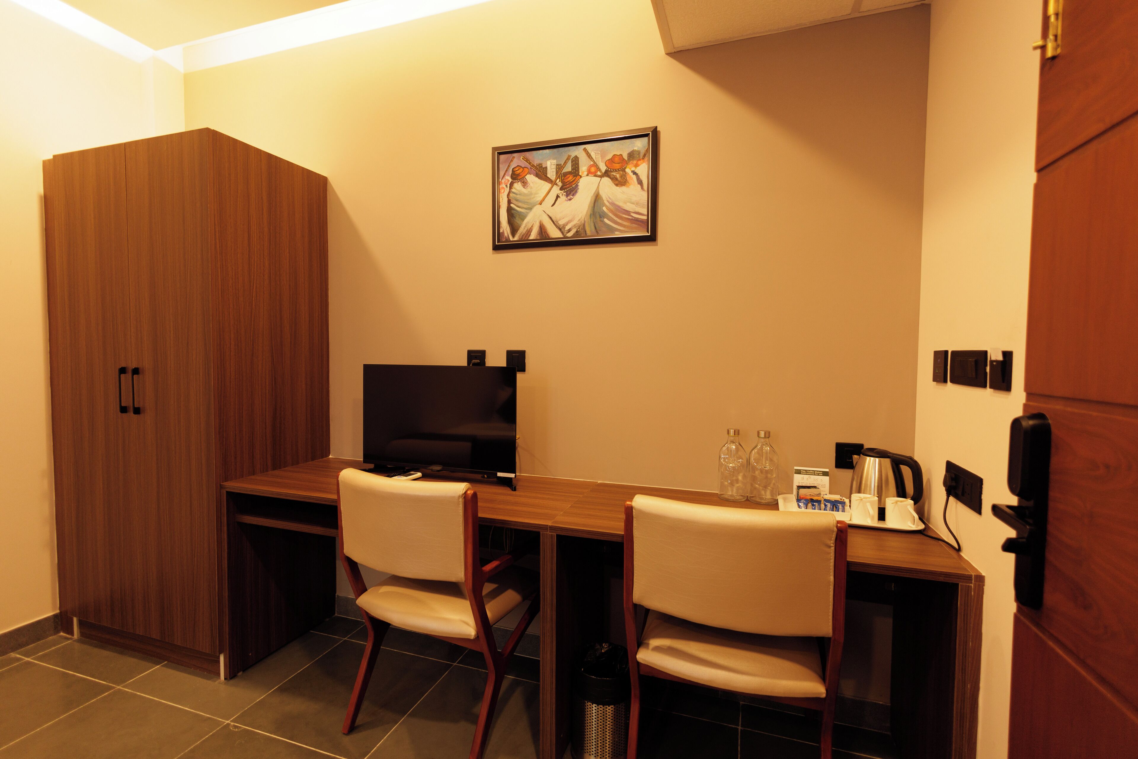 Quarto de hotel compacto com escrivaninha de madeira, duas cadeiras, televisão, pequena mesa com cafeteira e lixeira.