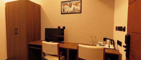 Quarto de hotel compacto com escrivaninha de madeira, duas cadeiras, televisão, pequena mesa com cafeteira e lixeira.