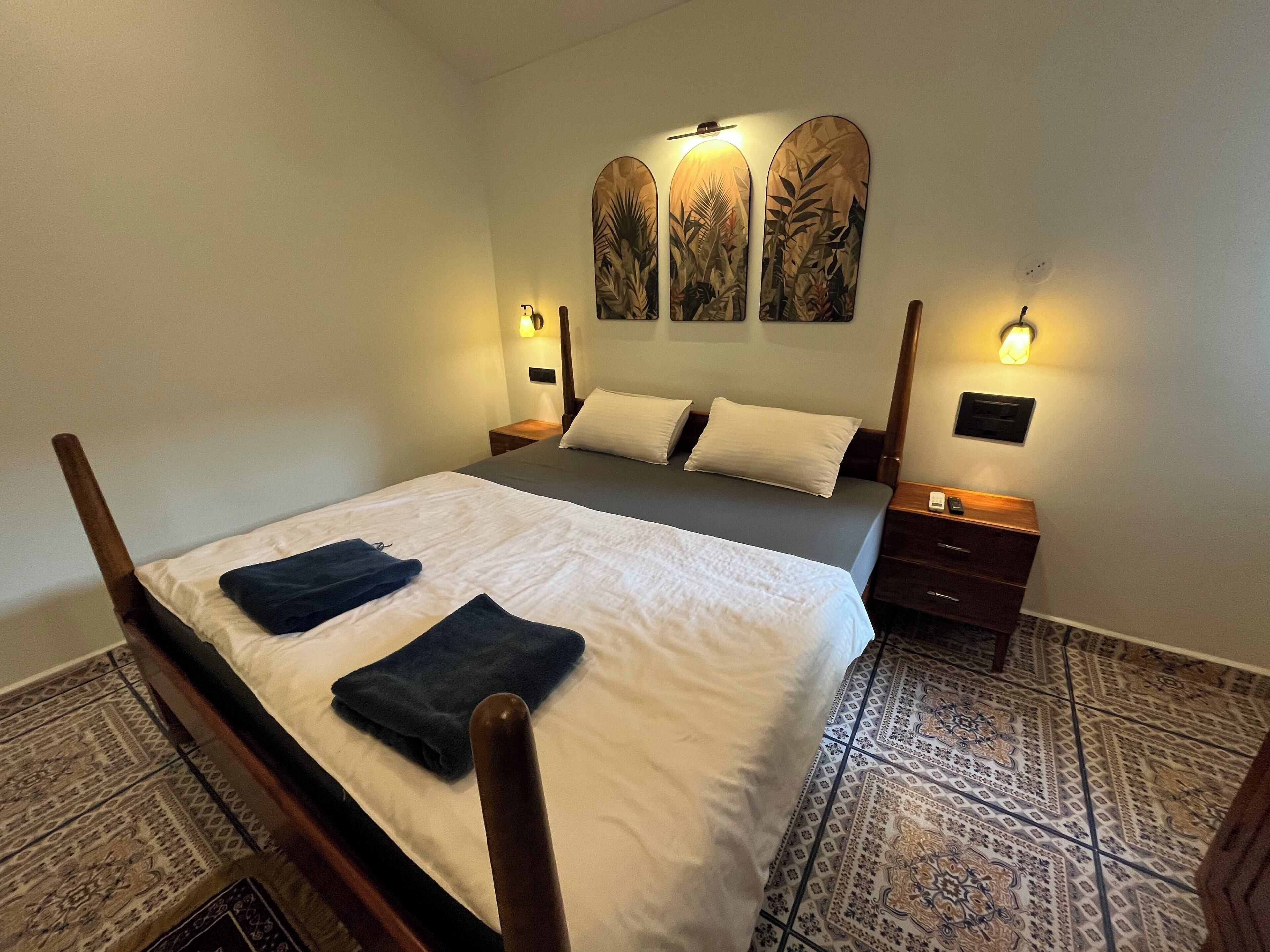 Cama bem arrumada com edredom cinza e branco, dois travesseiros e duas toalhas dobradas. Há duas mesas de cabeceira com abajures, uma obra de arte na parede e piso com ladrilhos estampados.