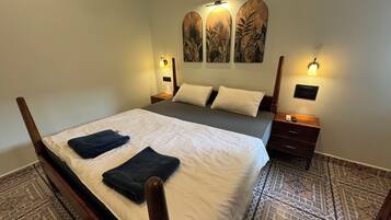 Cama bem arrumada com edredom cinza e branco, dois travesseiros e duas toalhas dobradas. Há duas mesas de cabeceira com abajures, uma obra de arte na parede e piso com ladrilhos estampados.