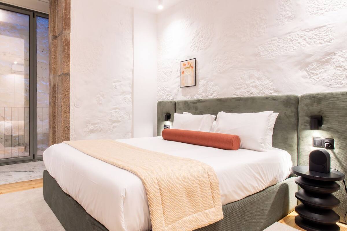 Quarto com cama, mesa de cabeceira, luminária na parede e vista por uma porta de vidro.