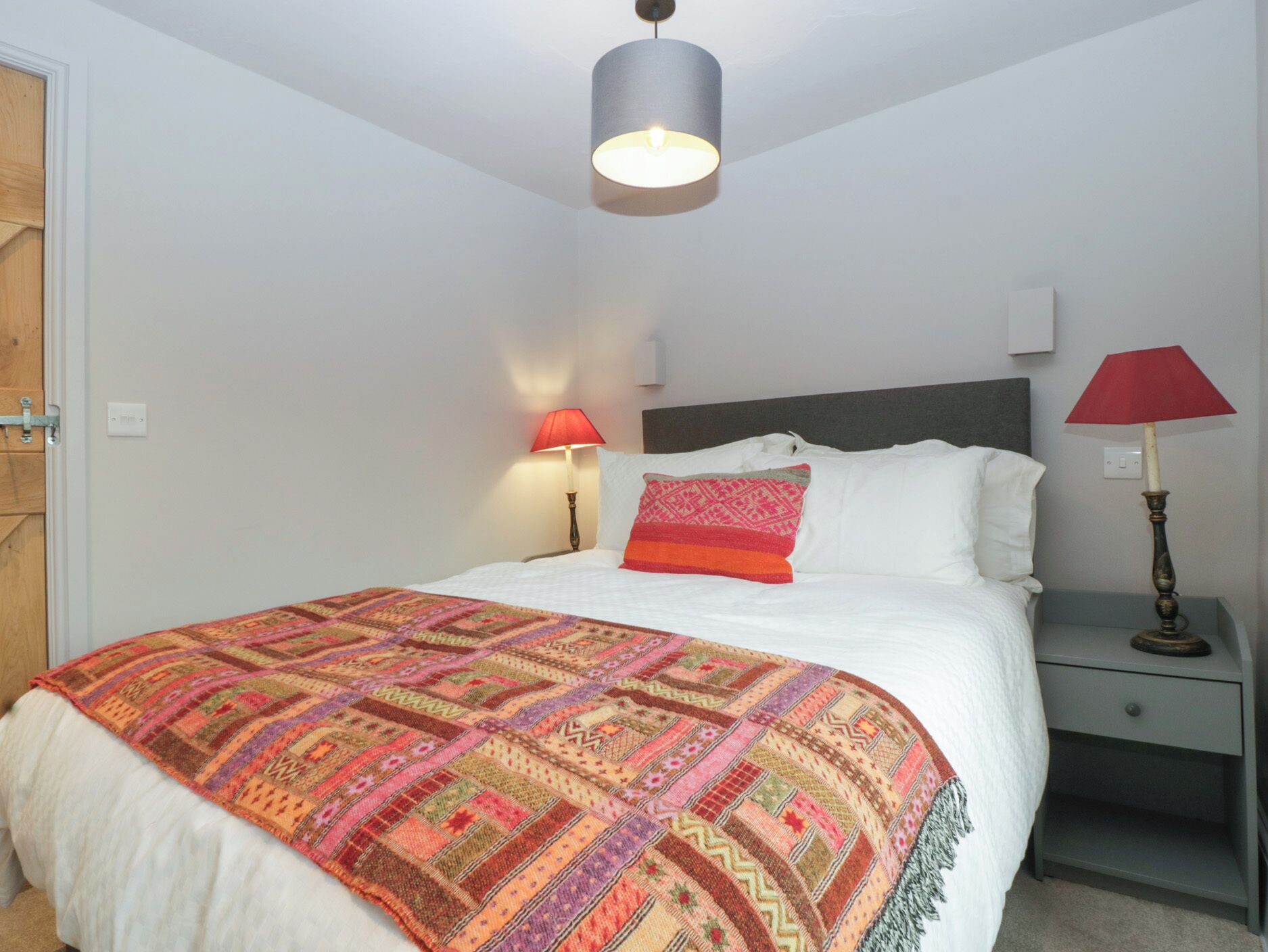 Cama bem arrumada com um cobertor colorido, duas luminárias de cabeceira e um lustre no teto.