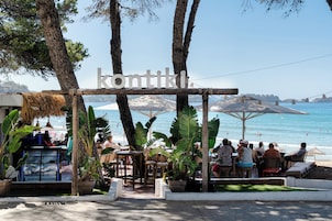 Um restaurante à beira-mar com mesas ao ar livre, telhado de palha e vista para o mar.