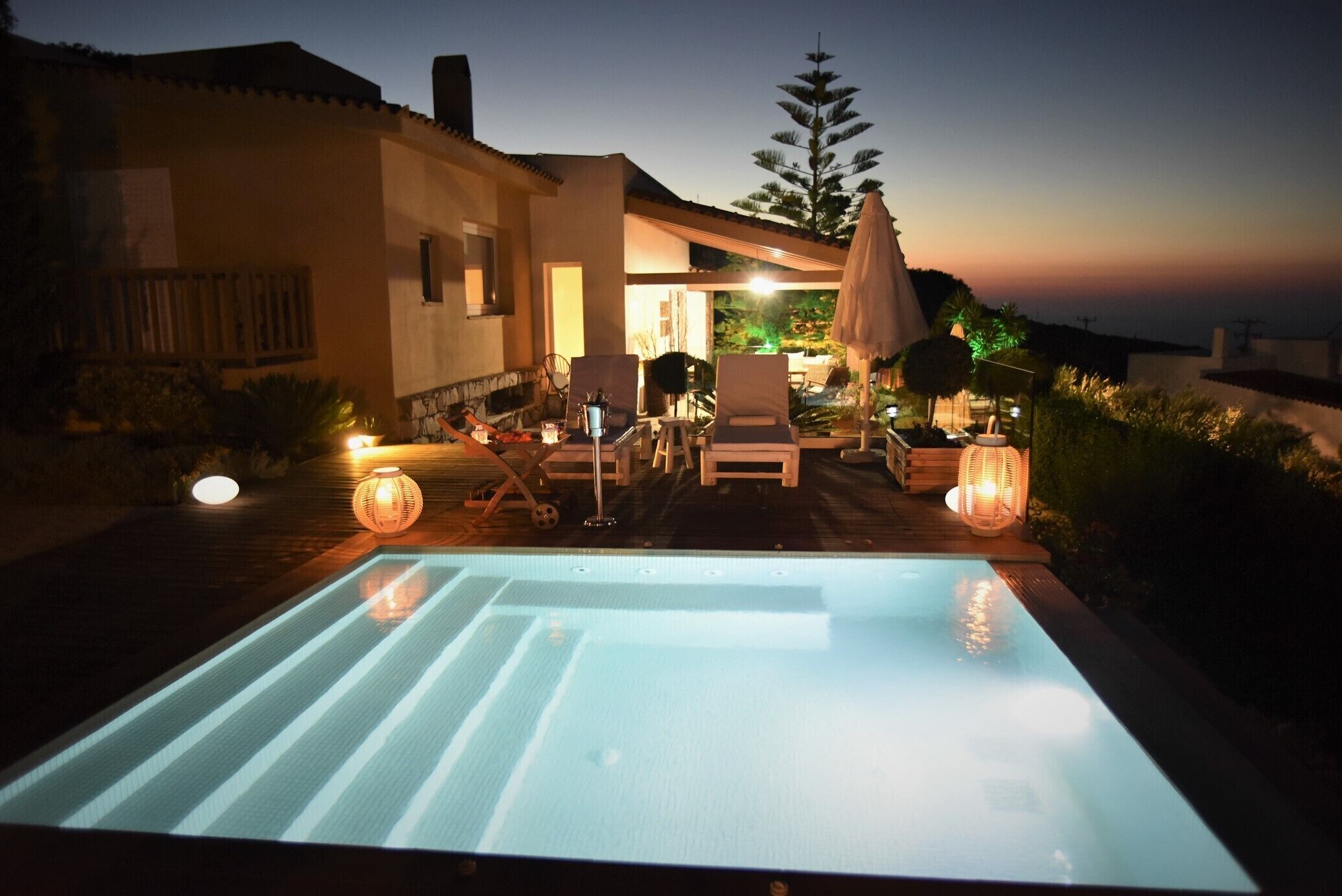 Uma área de piscina com espreguiçadeiras e banheira de hidromassagem, rodeada por um terraço com iluminação ambiente.