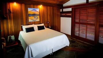 Quarto com uma cama grande, painéis de madeira nas paredes, um quadro de uma paisagem montanhosa e uma porta de correr com persianas horizontais.