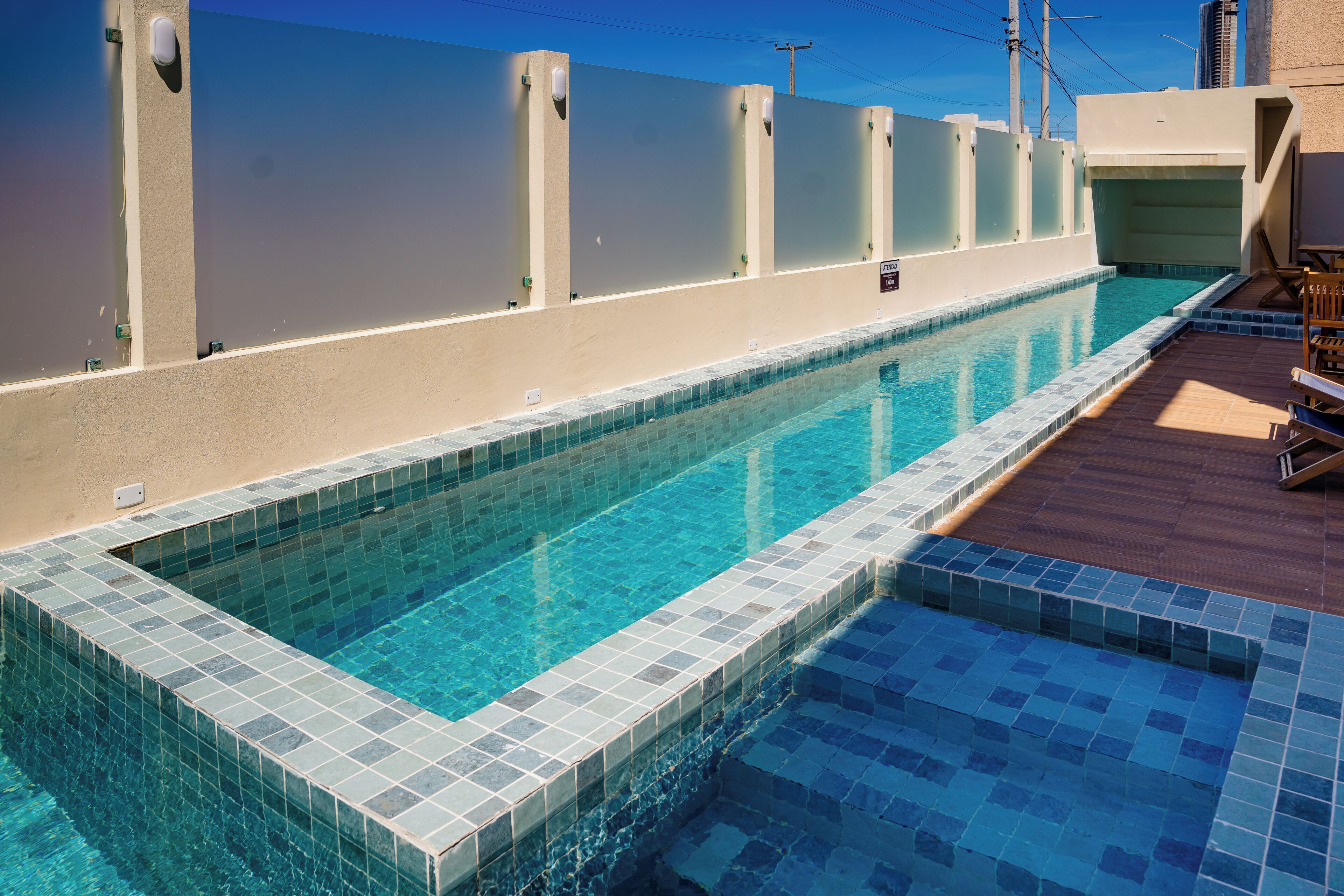 Piscina retangular com água azul cristalina, bordas azulejadas e área de deck de madeira com cadeiras.