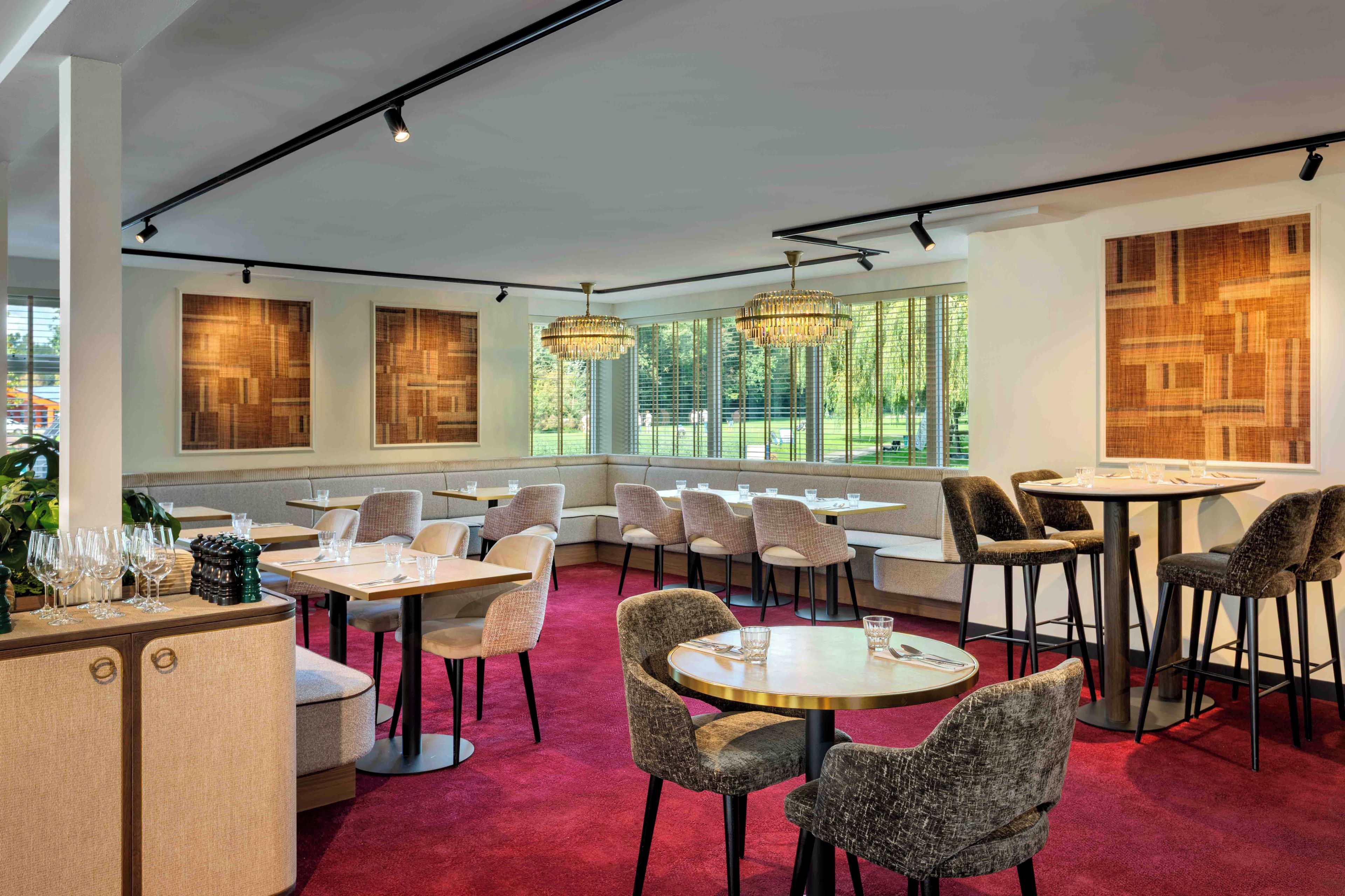 Restaurante com mesas redondas e assentos acolchoados, amplas janelas com vista para o verde e painéis decorativos nas paredes.