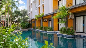 Piscina com bordas azulejadas, rodeada por vegetação e um edifício com varandas.