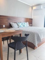 Quarto com cabeceira de madeira, cama com colcha cinza, duas almofadas estampadas, mesa de cabeceira com um jogo americano de palha e uma cadeira preta.