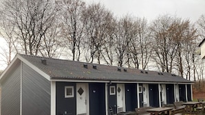 Uma fileira de cabanas térreas com portas numeradas, uma mesa de piquenique e bancos em uma área arborizada.