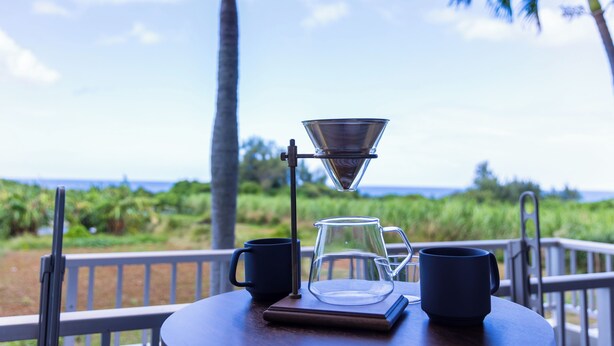 Uma mesa com uma cafeteira, duas canecas e um jarro de vidro numa varanda com vista para o oceano e vegetação.