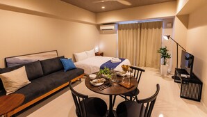 Quarto de hotel moderno com cama, mesa de jantar com cadeiras, sofá, ventilador de teto, ar condicionado, luminária e planta em vaso.