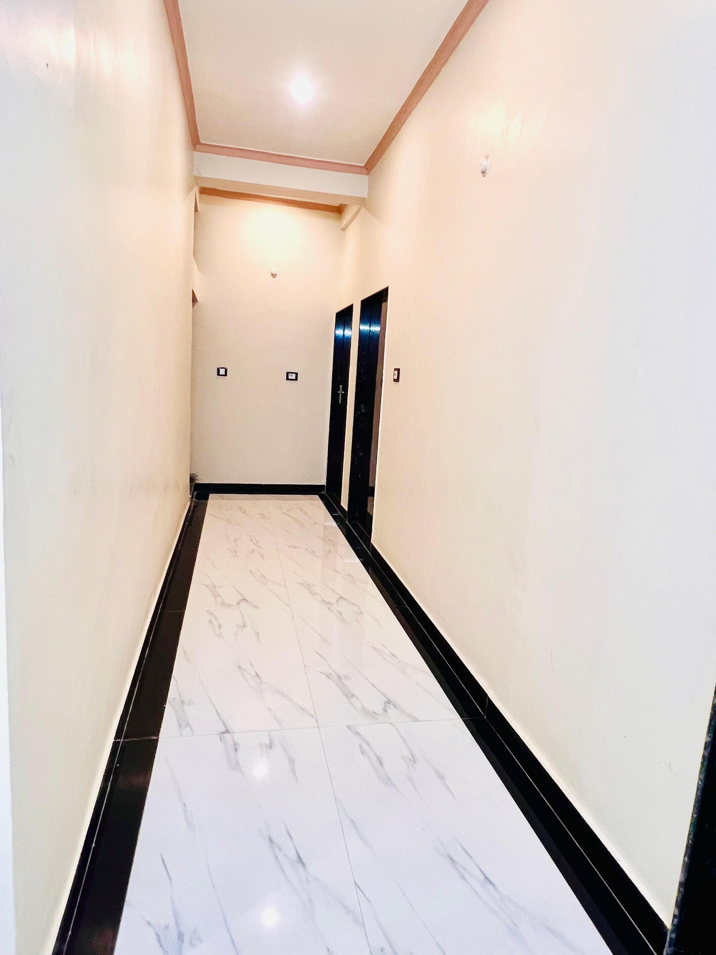 Corredor com piso de mármore, paredes brancas e detalhes em preto. Há duas portas com molduras pretas e um símbolo de cruz na parede.