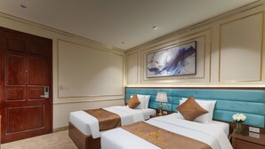 Quarto de hotel com duas camas, uma porta de madeira grande, cabeceira azul e um quadro na parede.