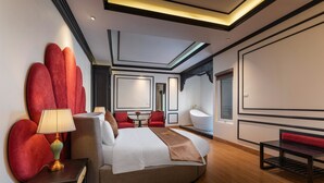 Quarto de hotel moderno com cama curva, cadeiras estofadas em vermelho, uma mesa redonda pequena e um espelho grande.