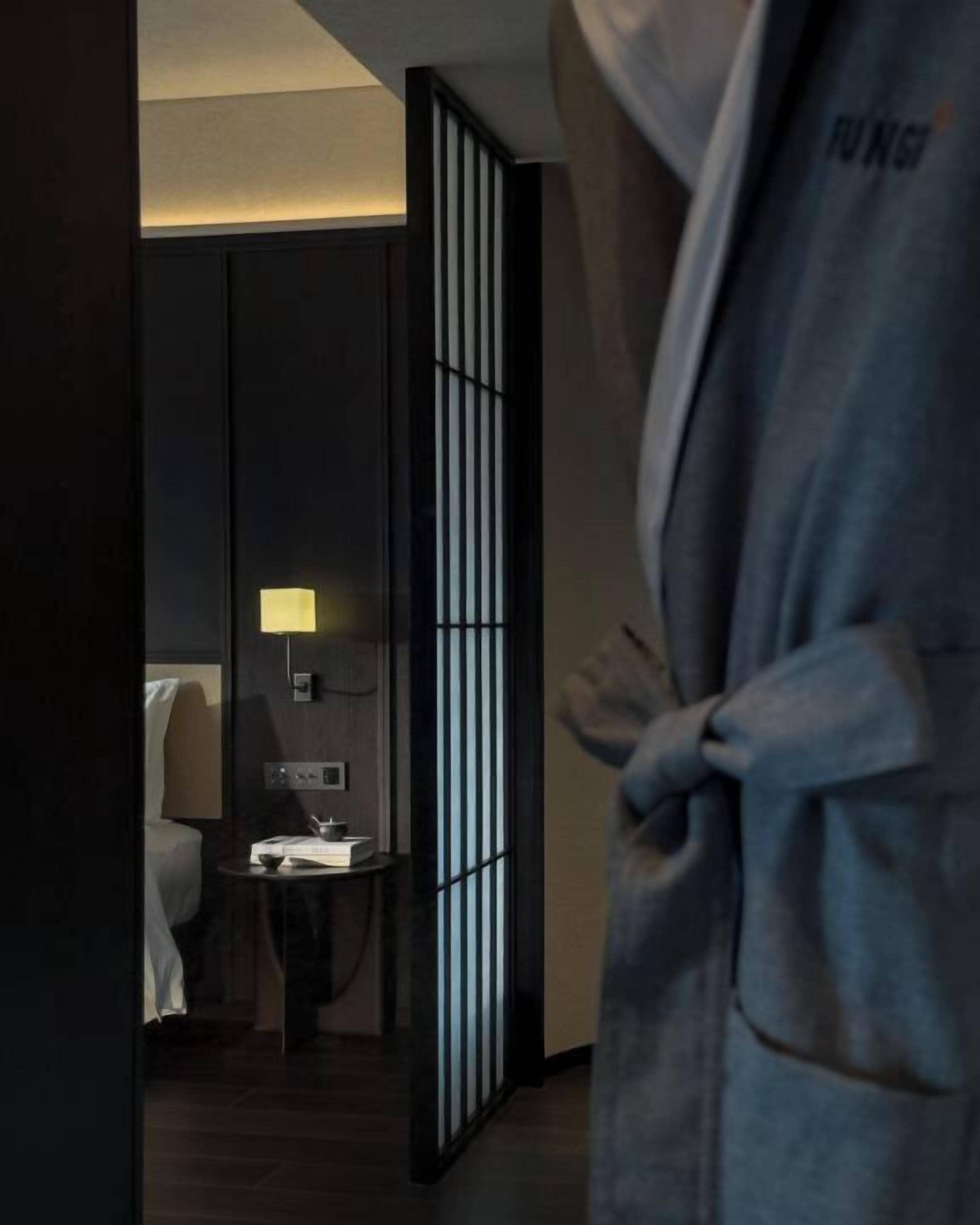 Quarto de hotel com cama, mesa de cabeceira, abajur e porta deslizante que dá para outro cômodo.
