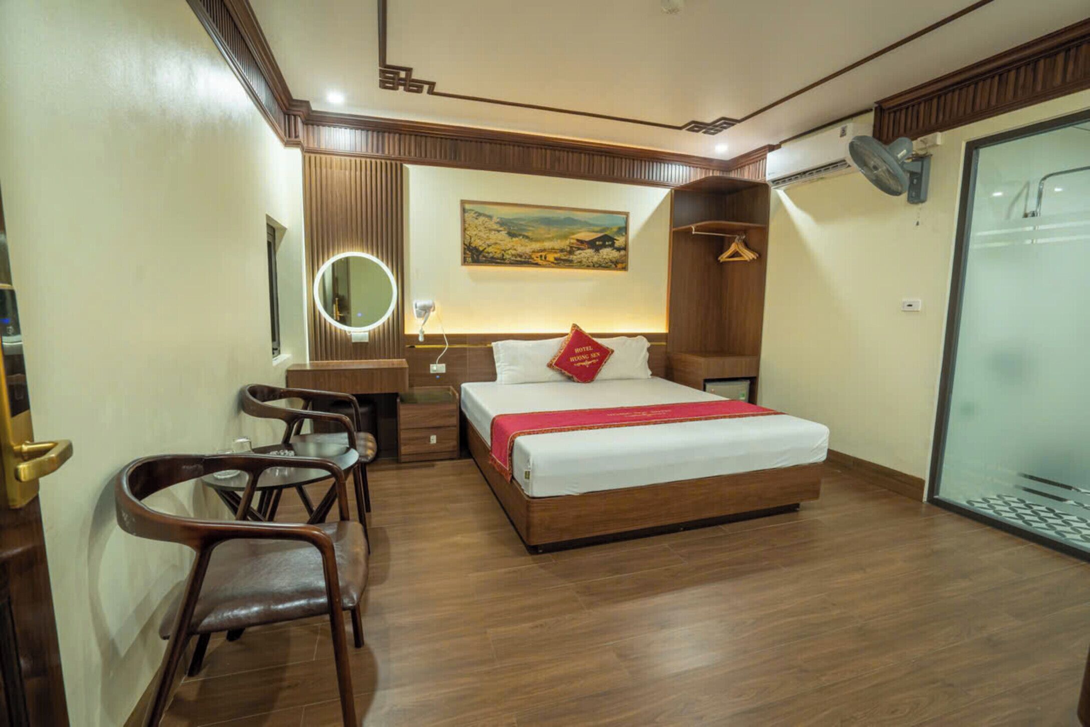 Quarto de hotel com cama de madeira, uma escrivaninha com cadeira, um espelho e um quadro na parede.