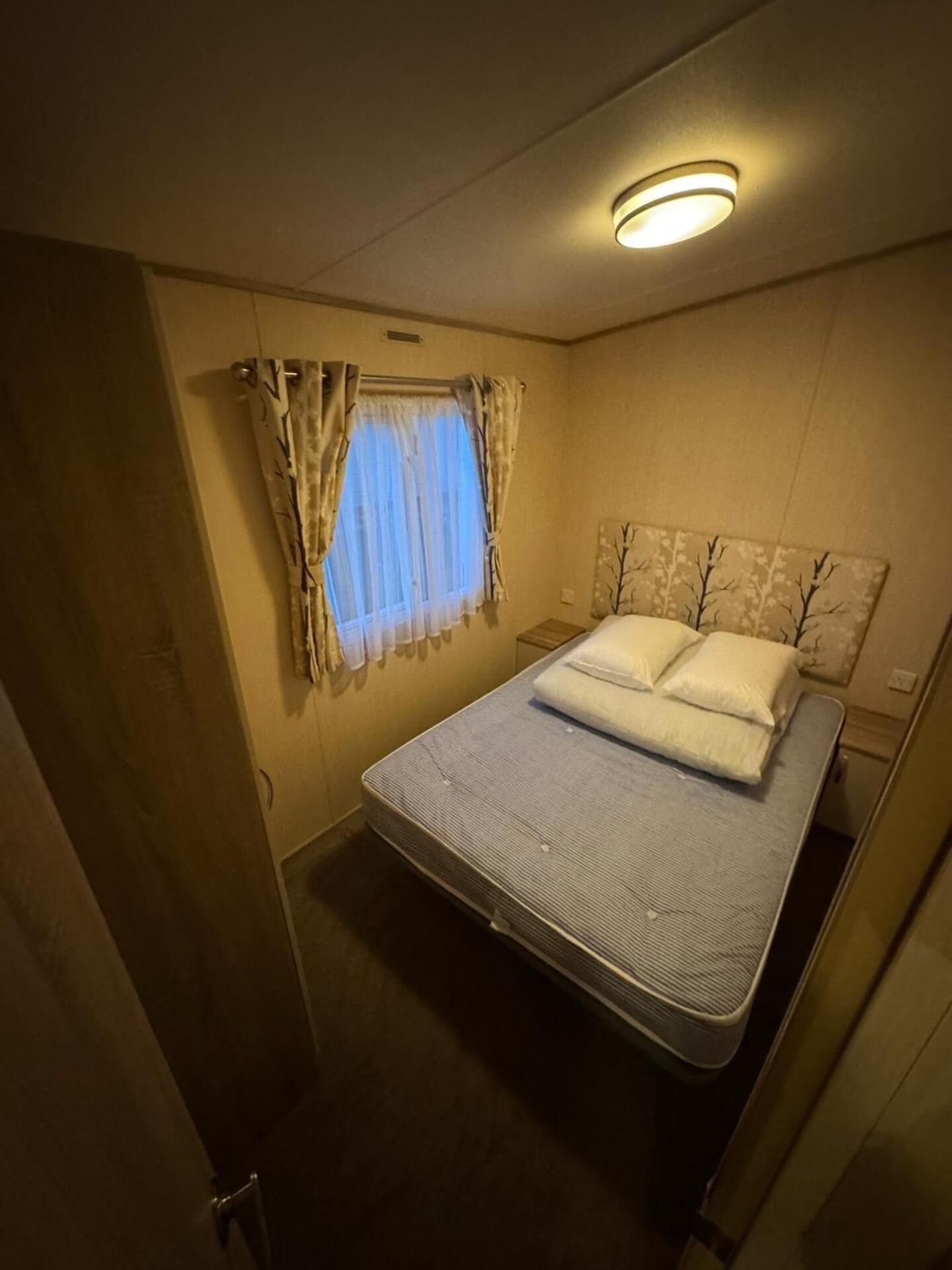 Quarto compacto com cama, janela com cortinas, cabeceira com padrão de árvore e luminária no teto.