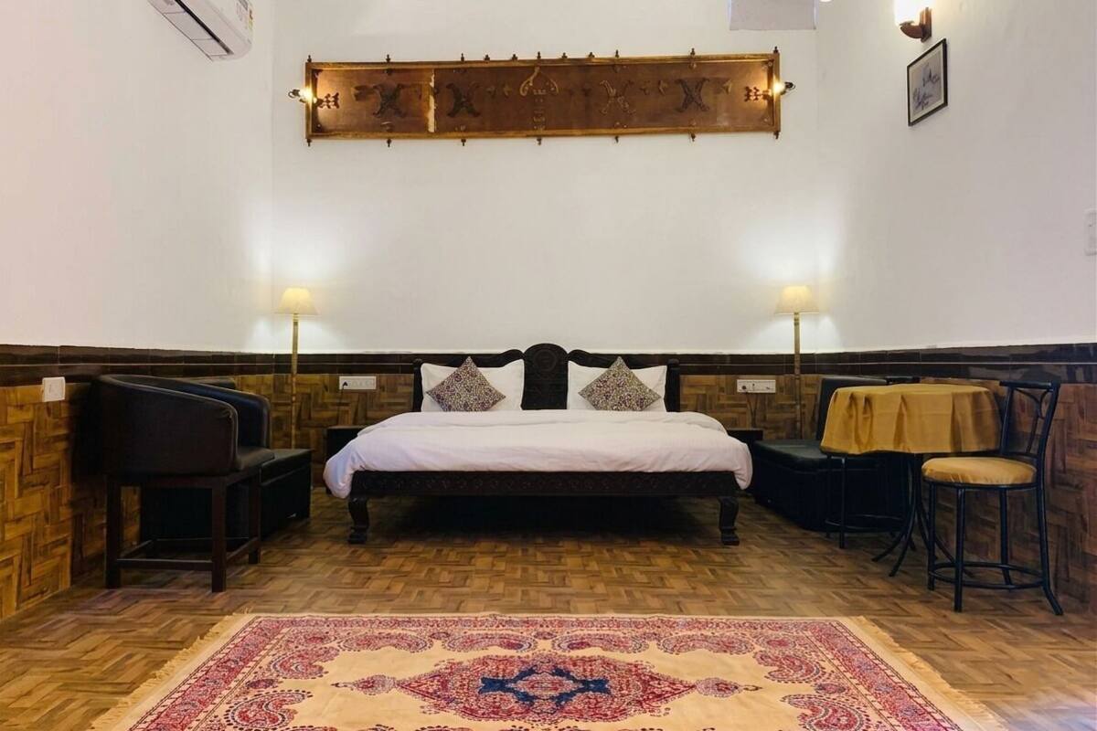 Quarto com cabeceira de madeira, cama grande, duas luminárias de cabeceira, piso de madeira e tapete estampado.