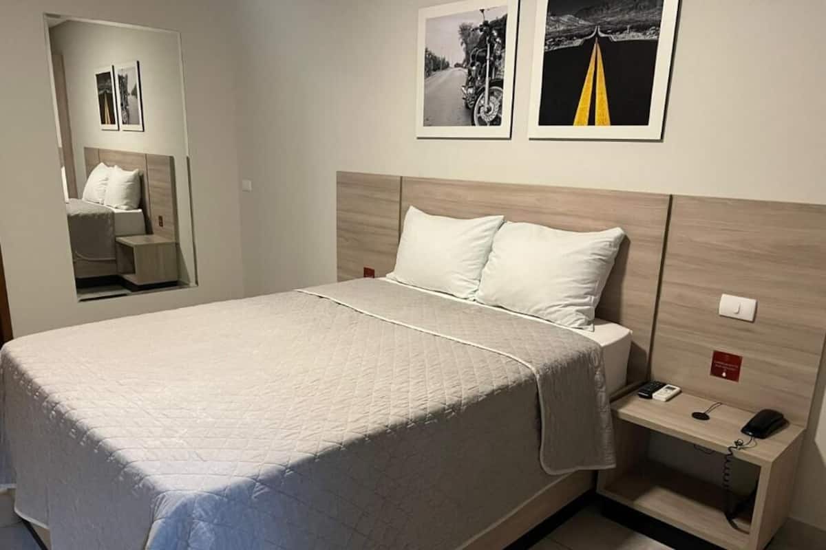 Uma cama bem arrumada com manta acolchoada, uma mesa de cabeceira com telefone e abajur, e quadros nas paredes.