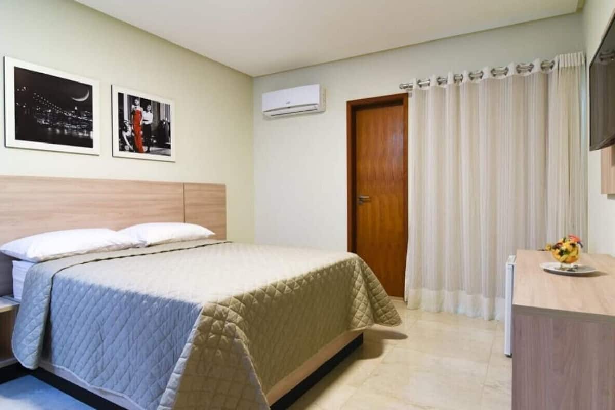 Quarto com cama, cabeceira de madeira, escrivaninha com um vaso e porta com cortinas.