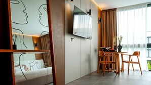Quarto moderno com cama, televisão fixada na parede, área de jantar com cadeiras e uma janela grande com cortinas.